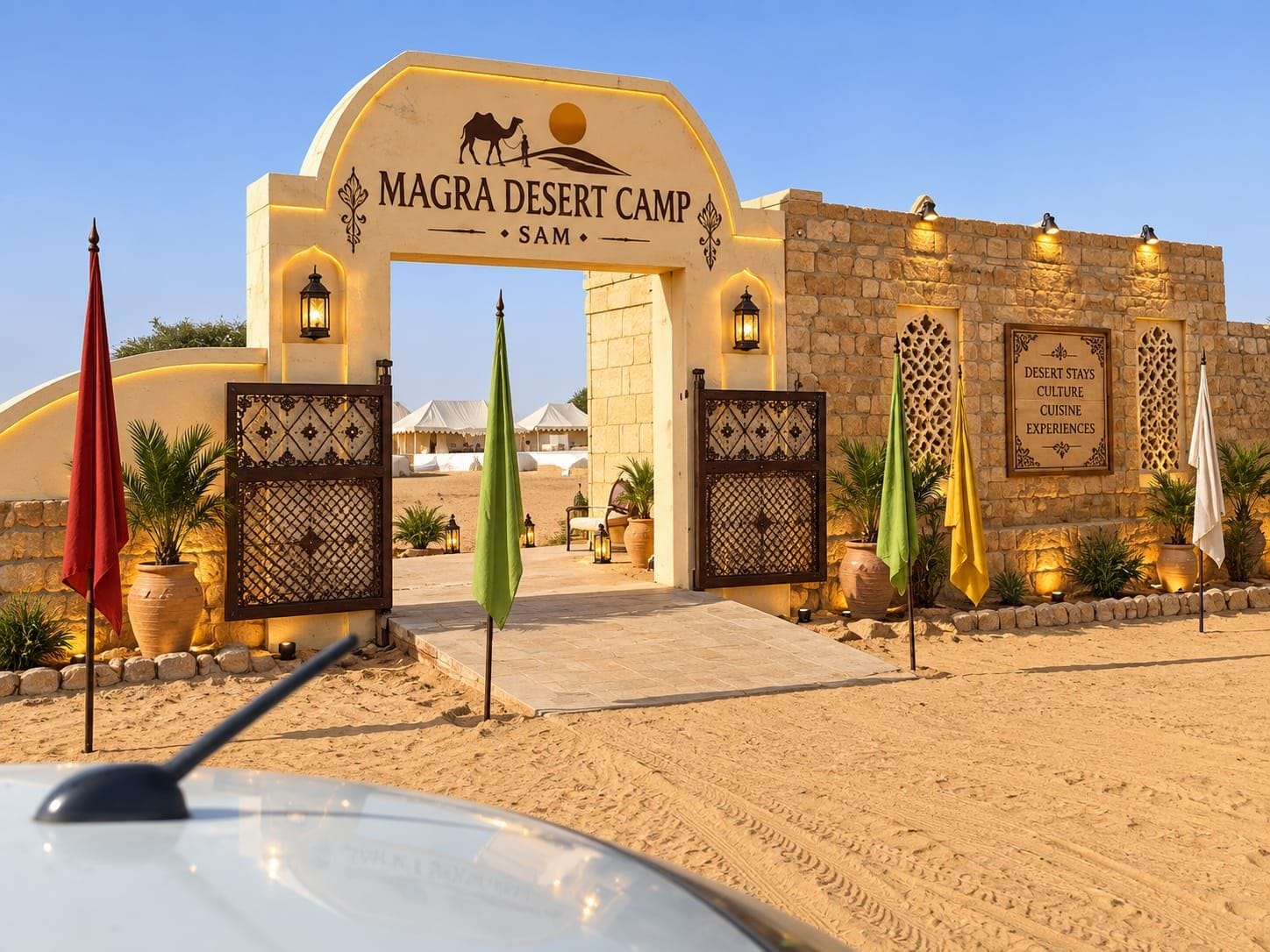 A entrada do Magra Desert Camp, com silhueta de camelo e um sol gráfico, uma parede de pedra com a placa 'Desert Stays Culture' e um caminho pavimentado que leva às tendas.