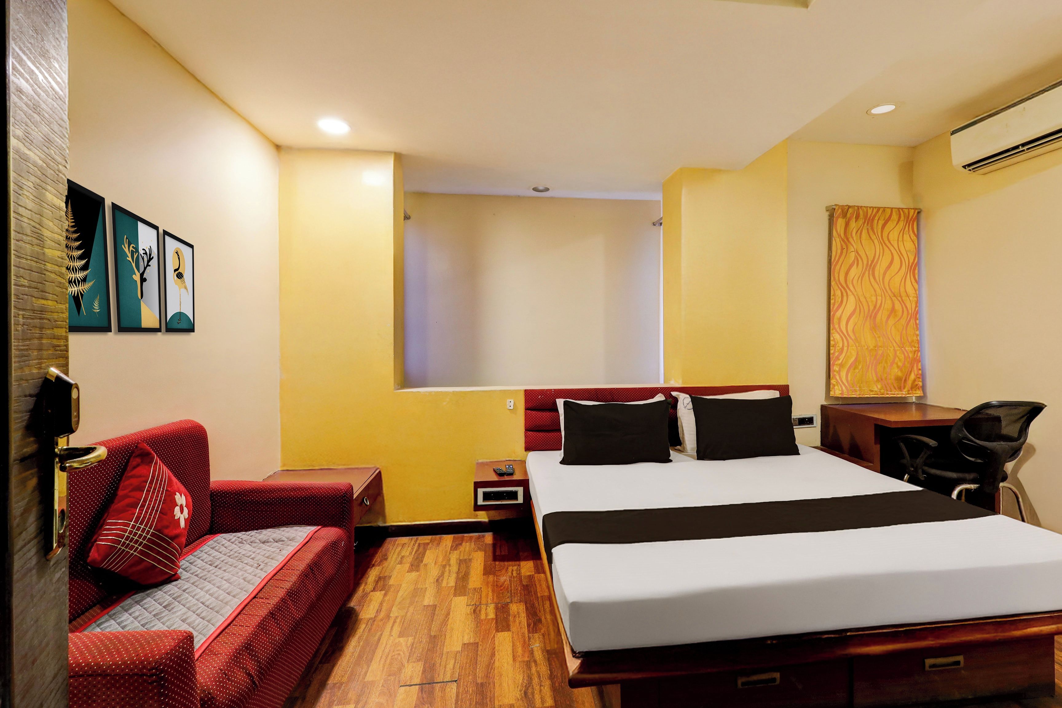 Quarto de hotel com cama, escrivaninha, cadeira, sofá vermelho e obras de arte na parede.