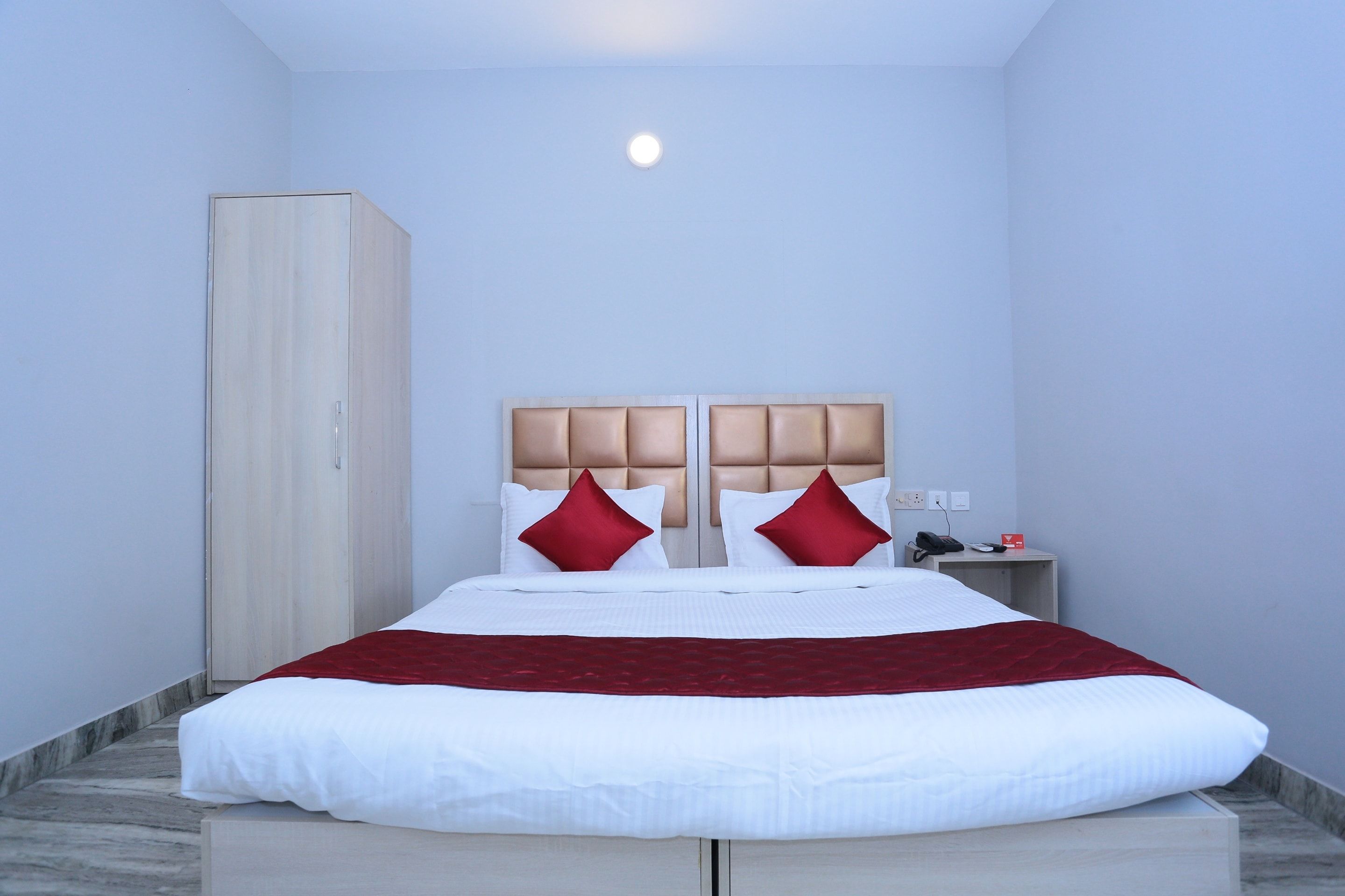 Quarto de hotel com uma cama, guarda-roupa, mesa de cabeceira com telefone e luminária no teto.