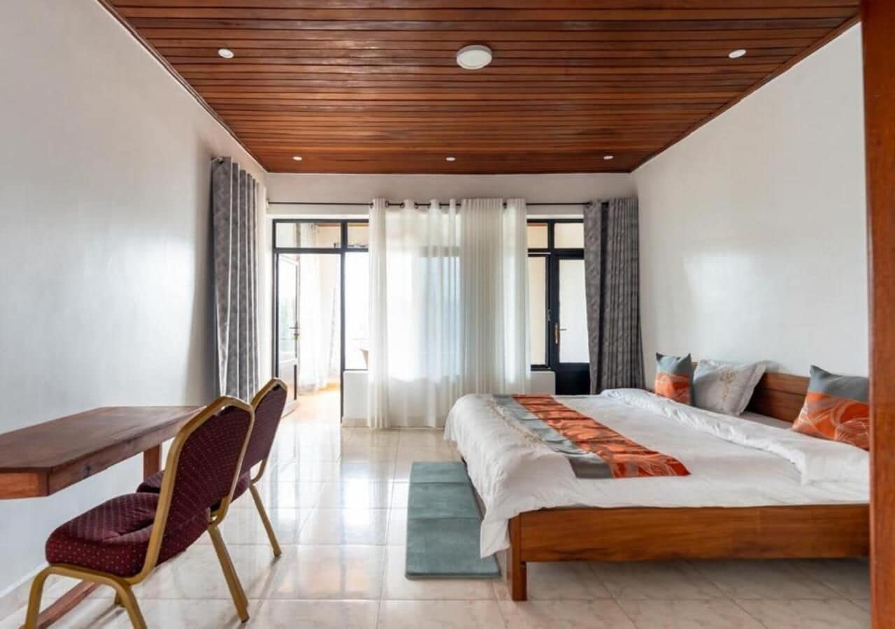 Quarto moderno com teto de madeira, uma cama grande com várias almofadas, uma escrivaninha com cadeiras e vista através de uma janela ampla.