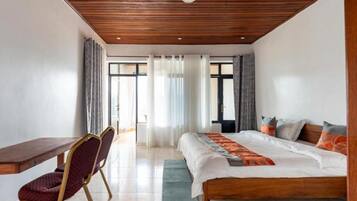 Quarto moderno com teto de madeira, uma cama grande com várias almofadas, uma escrivaninha com cadeiras e vista através de uma janela ampla.