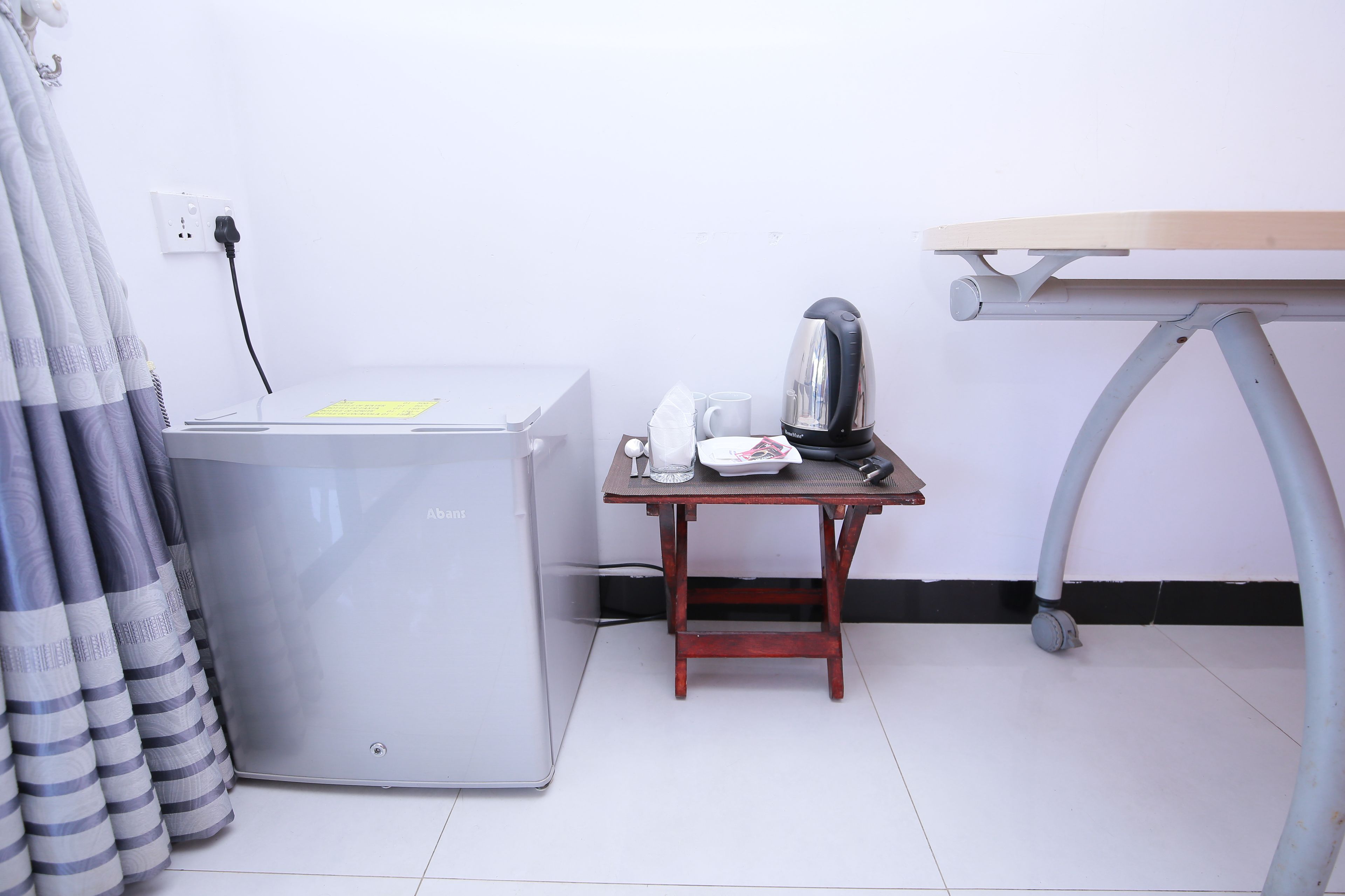 Uma cozinha compacta com geladeira, uma mesa pequena com chaleira e uma tomada na parede.