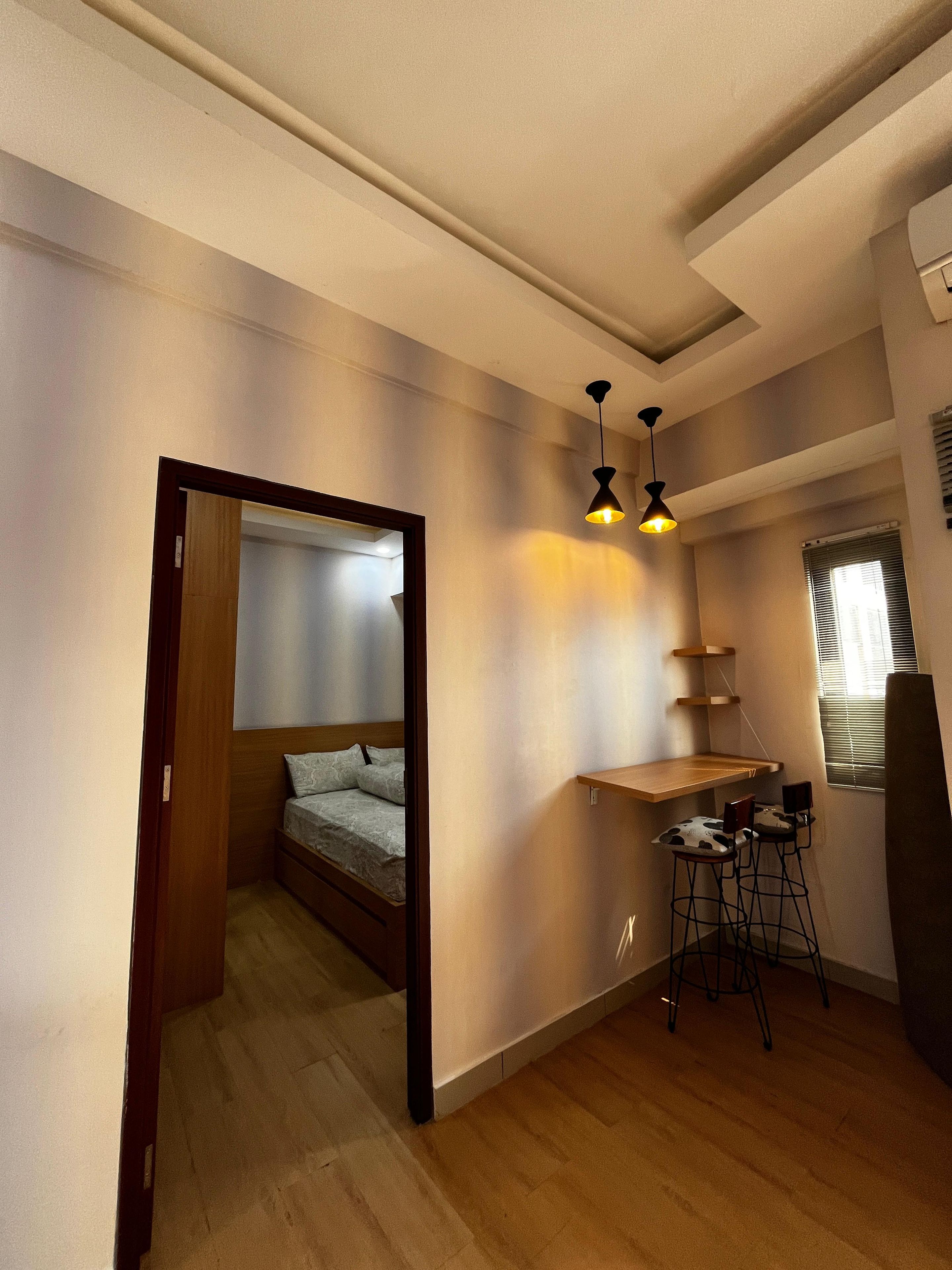 Quarto compacto com cama, escrivaninha com banquinhos, luminária pendente e janela com persianas.