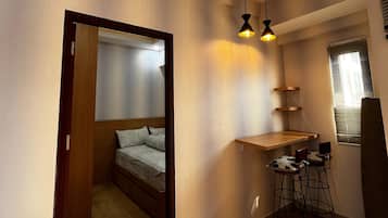 Quarto compacto com cama, escrivaninha com banquinhos, luminária pendente e janela com persianas.