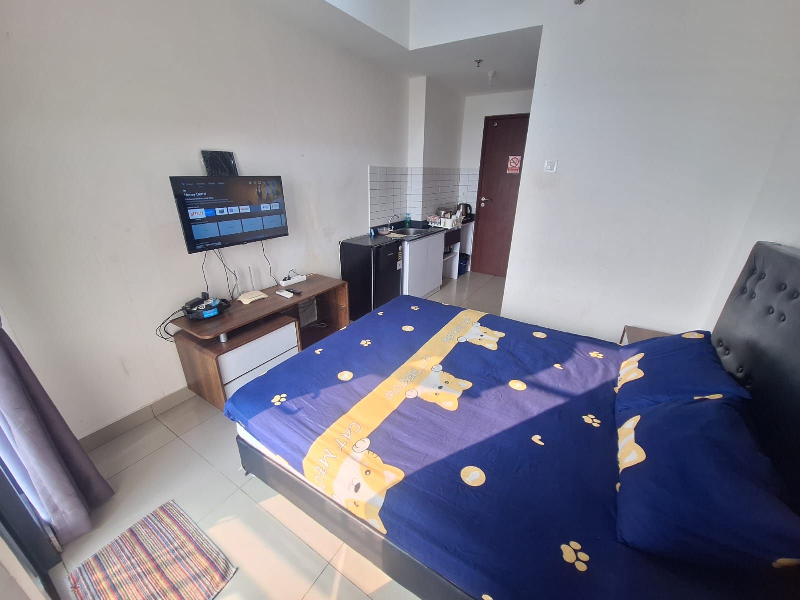Quarto de hotel com cama, escrivaninha com computador, televisão e cozinha compacta.