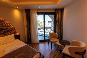 Quarto de hotel com uma cama, duas cadeiras, uma mesinha e vista para uma piscina e edifícios lá fora.