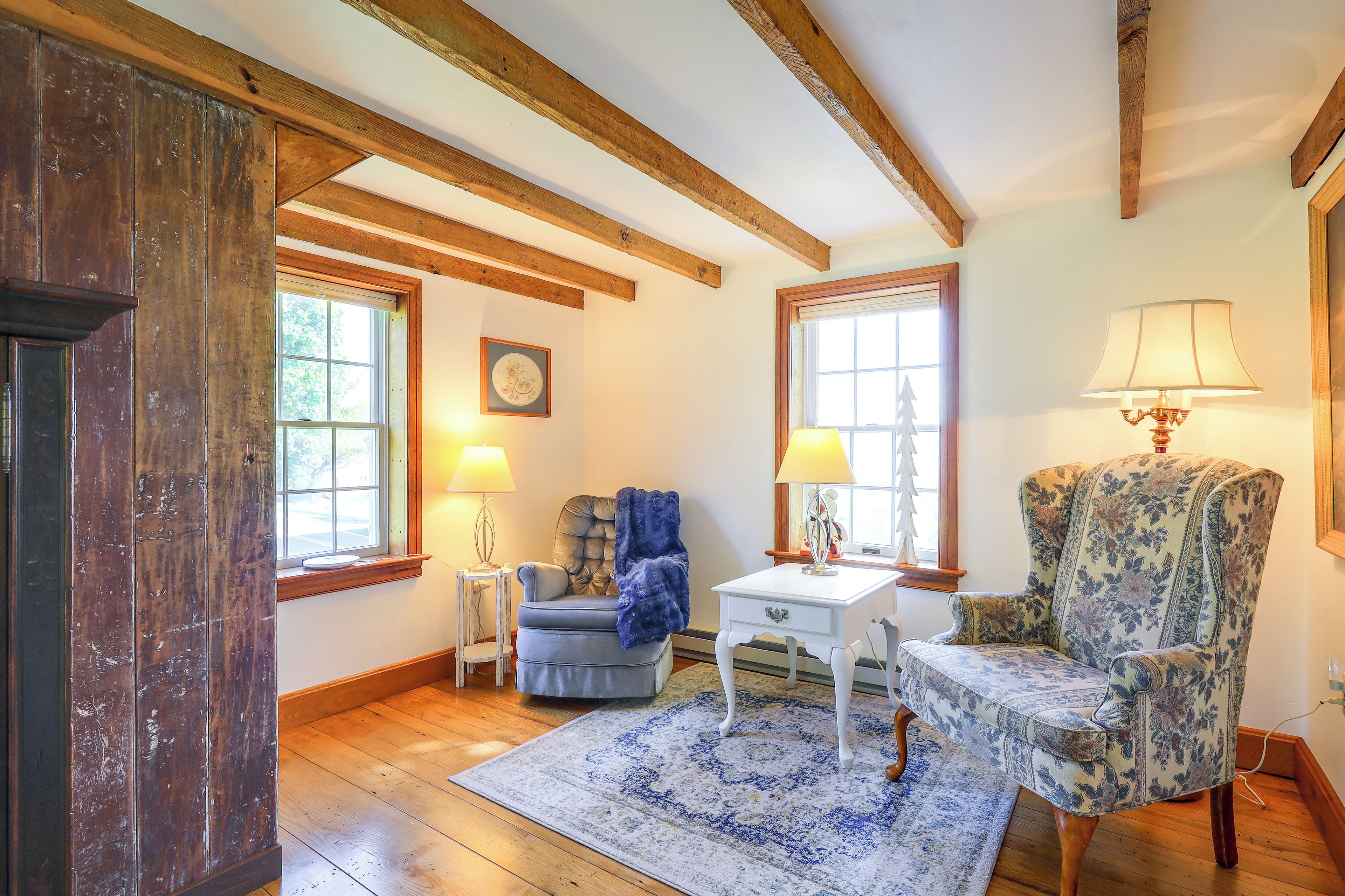 Quarto com vigas de madeira, uma poltrona azul, uma mesa branca e outra poltrona estampada.
