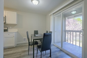 Cozinha compacta com armários brancos, micro-ondas e um frigorífico pequeno. Há uma mesa de jantar com duas cadeiras e um laptop sobre ela. O quarto possui uma grande porta de correr de vidro que dá para uma área externa com vista para árvores.