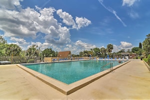 Piscina retangular com água azul cristalina, ladeada por um deck de concreto. Cadeiras de descanso estão dispostas ao longo da borda da piscina, e um edifício com telhado marrom é visível ao fundo.