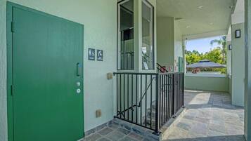 Área externa coberta com porta verde, grade de metal e vista para um estacionamento.
