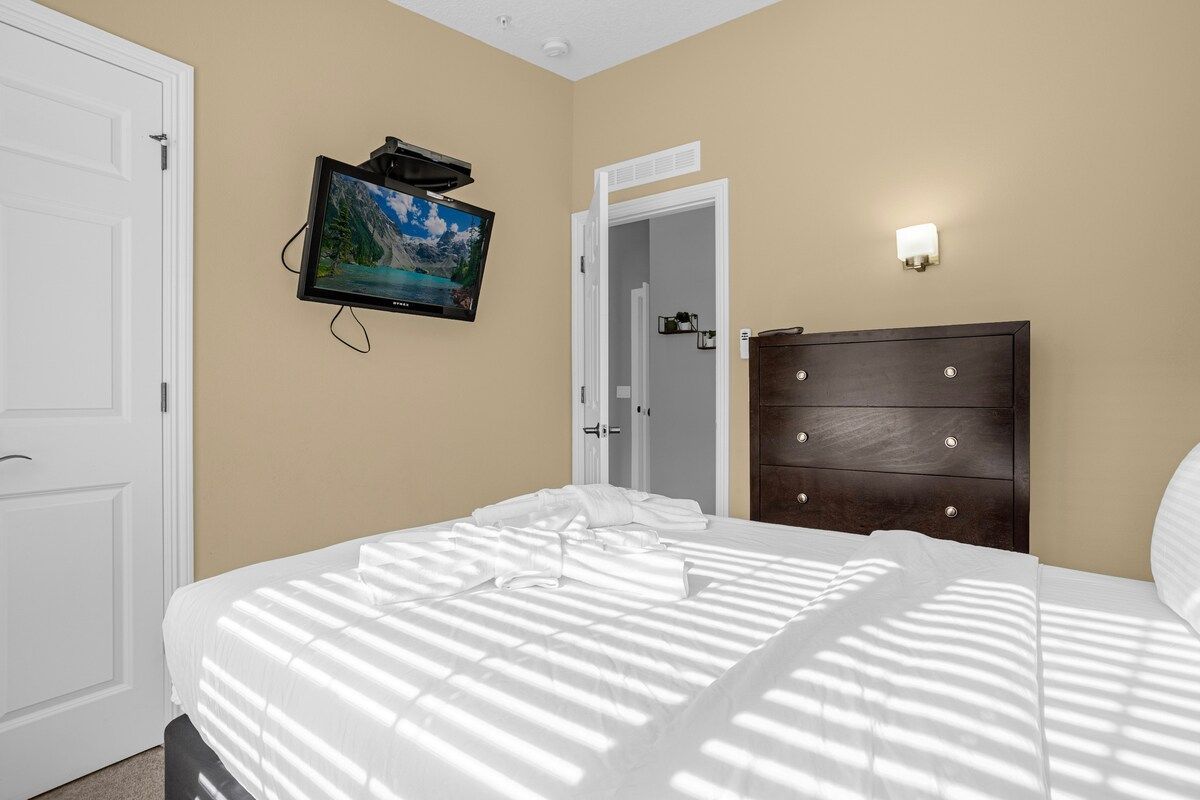 Quarto de hotel com cama, televisão presa à parede, penteadeira e porta que dá para outro espaço.