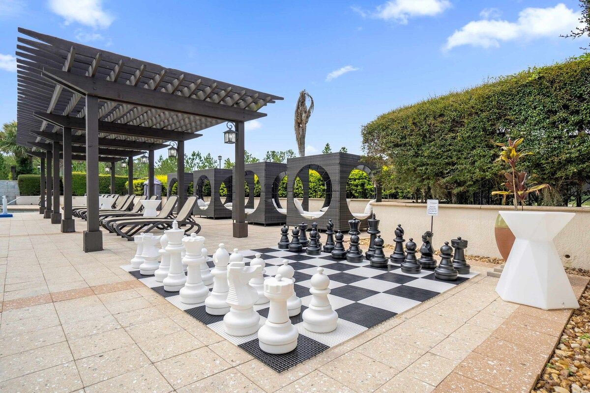 Tabuleiro de xadrez com peças grandes montado ao ar livre, cercado por cadeiras de descanso e uma estrutura de pérgola.