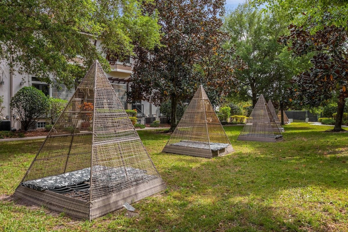 Áreas de estar ao ar livre com estrutura de metal e almofadas, cercadas por árvores e um edifício ao fundo.