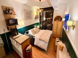 Quarto de hotel compacto com cama, escrivaninha, pequeno espaço de cozinha e guarda-roupa.