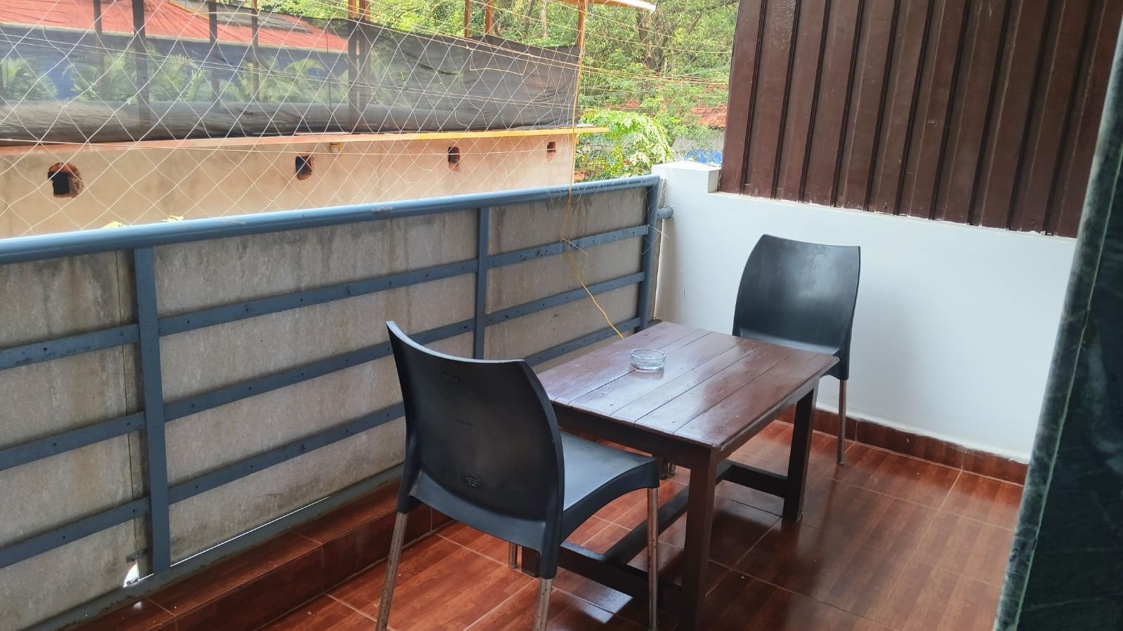 Uma varanda com mesa e duas cadeiras, guarda-corpo de metal e vista para o verde.