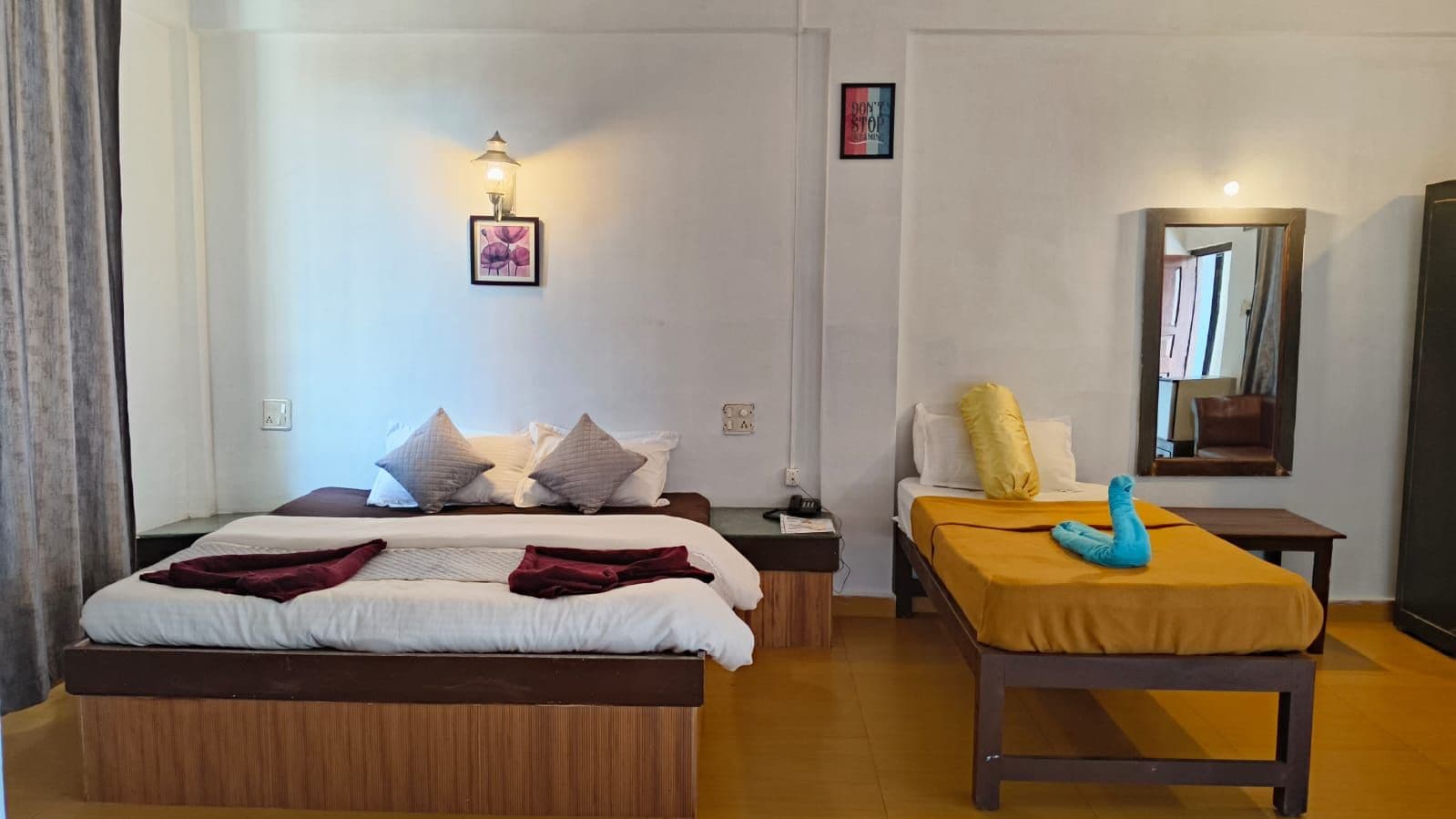 Quarto de hotel com uma cama grande, uma cama menor, uma mesa de cabeceira, um criado-mudo, um espelho e um aplique na parede.