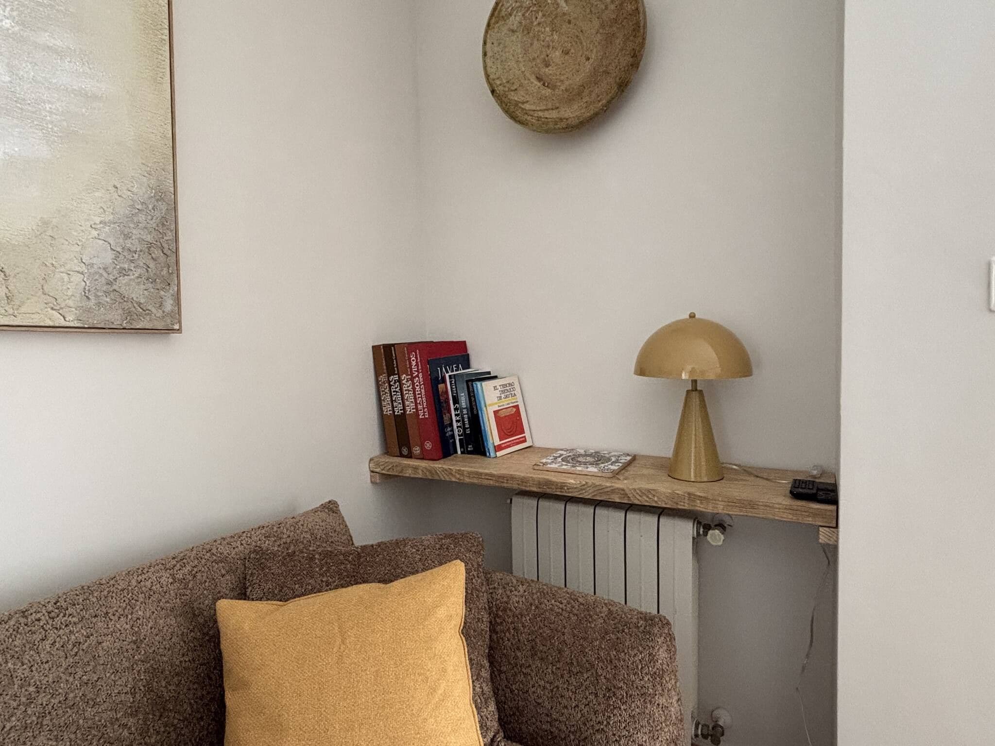 Uma sala de estar com um sofá marrom, um travesseiro amarelo, uma estante com livros, um abajur e um radiador.