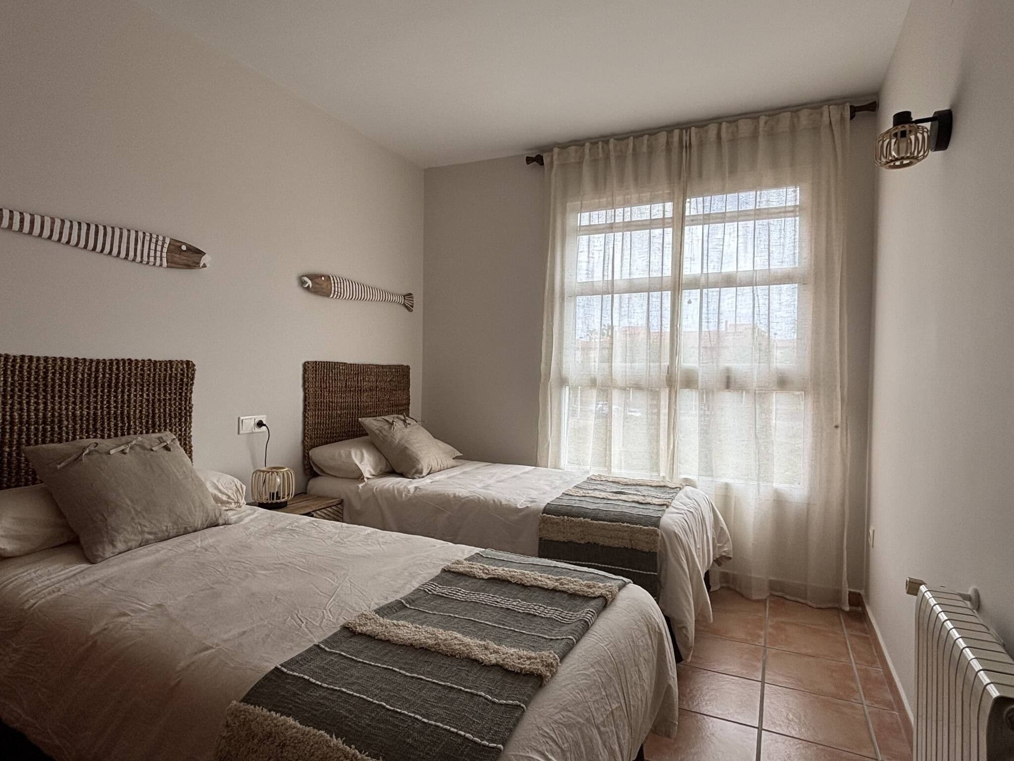 Quarto com duas camas, uma janela grande com cortinas leves e um radiador na parede.