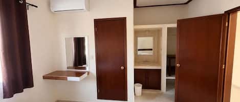 Quarto de hotel com cama, pequena escrivaninha, ar condicionado, espelho e banheiro com pia e espelho.
