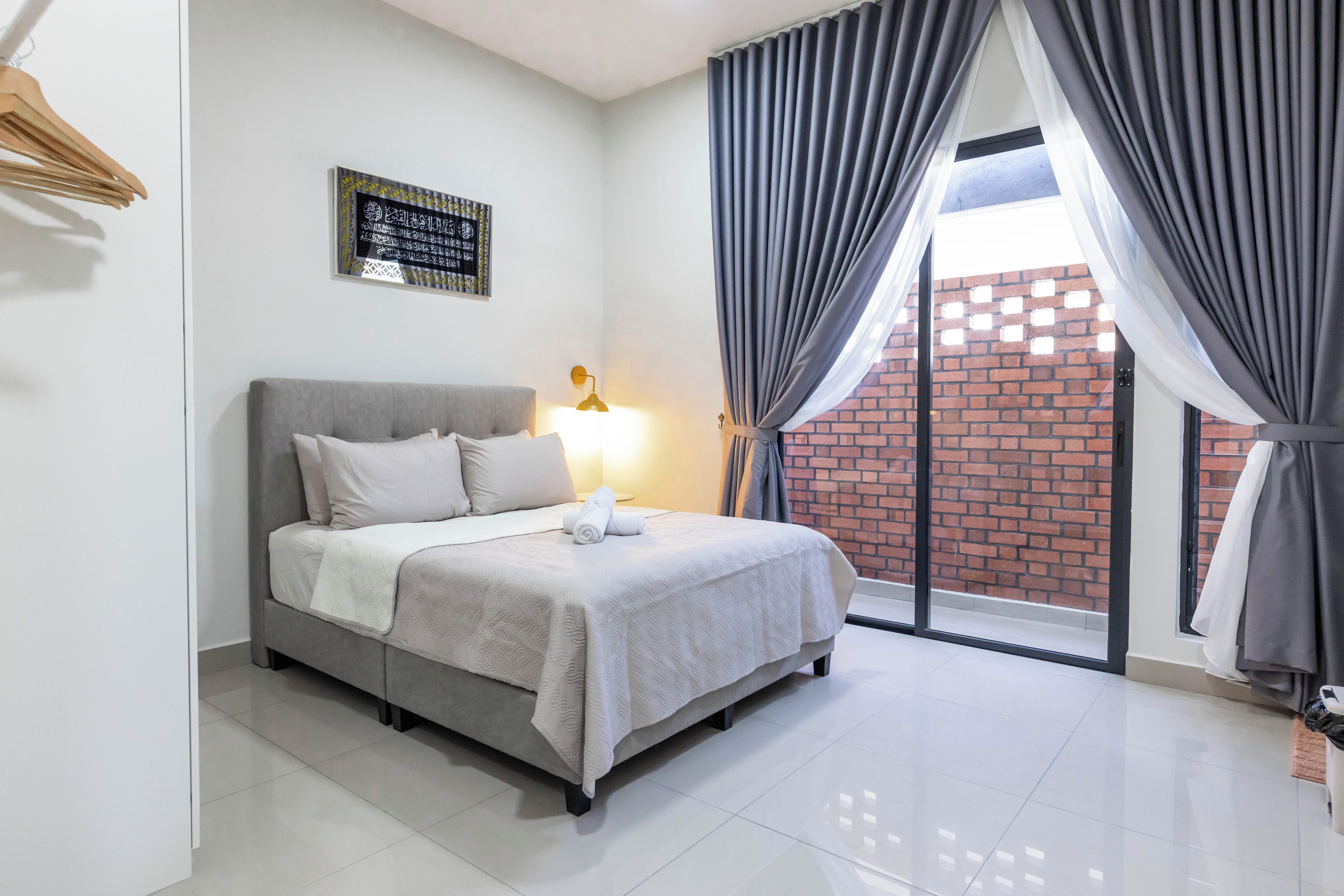 Quarto com cama, luminária na parede e vista para uma parede de tijolos através de uma janela com cortinas.