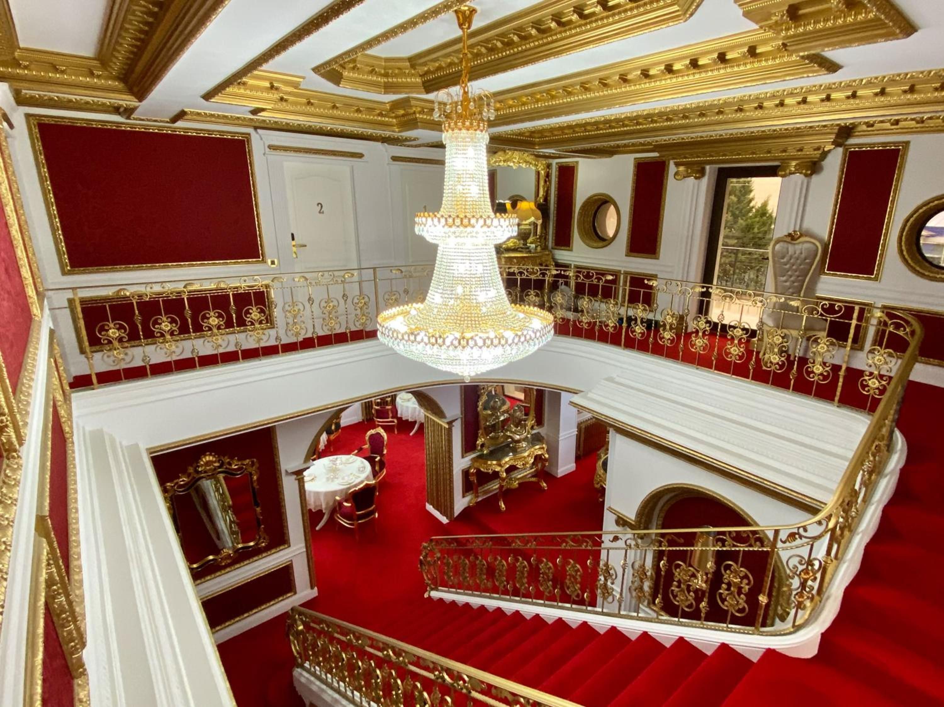 Um interior imponente com um lustre central, pisos cobertos por um tapete vermelho e detalhes em dourado ornamentado.