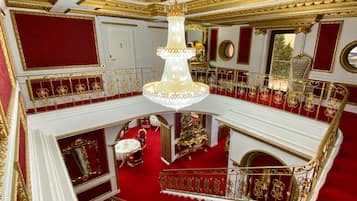 Um interior imponente com um lustre central, pisos cobertos por um tapete vermelho e detalhes em dourado ornamentado.