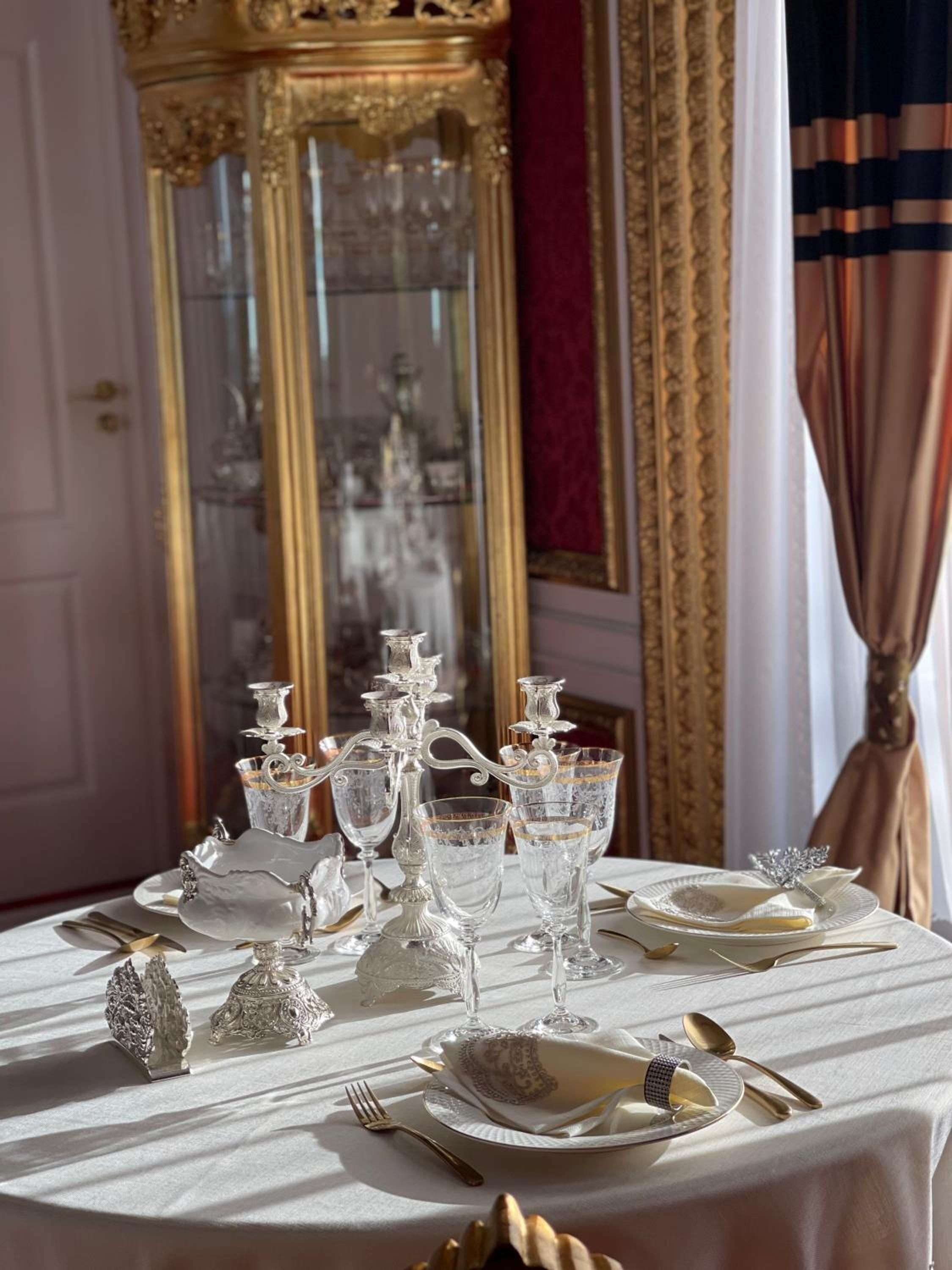 Mesa de jantar formal posta com talheres, pratos e copos, com um espelho e cortinas ao fundo.