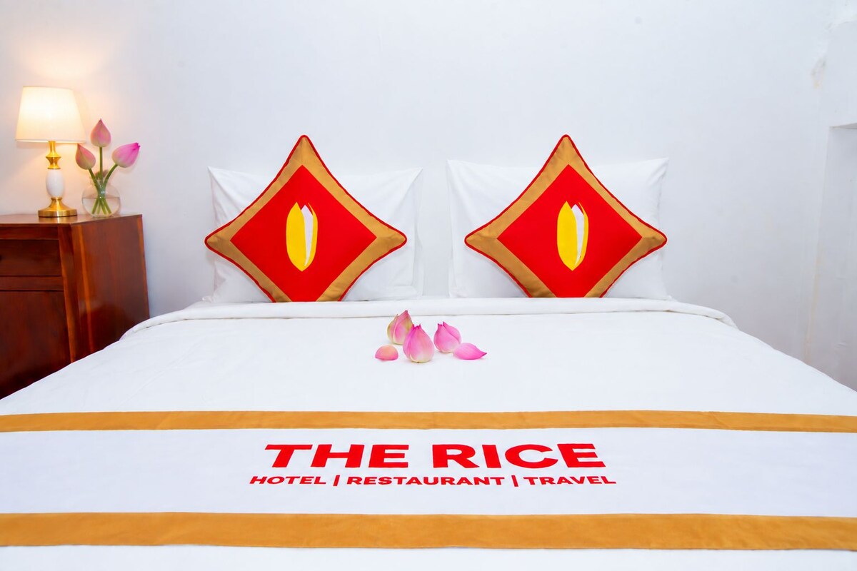 Quarto de hotel com cama, almofadas, mesa de cabeceira com luminária e sinalização indicando serviços como hotel, restaurante e viagens.