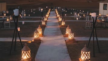 Um caminho no deserto iluminado por lanternas, levando a um edifício com mesas e cadeiras.