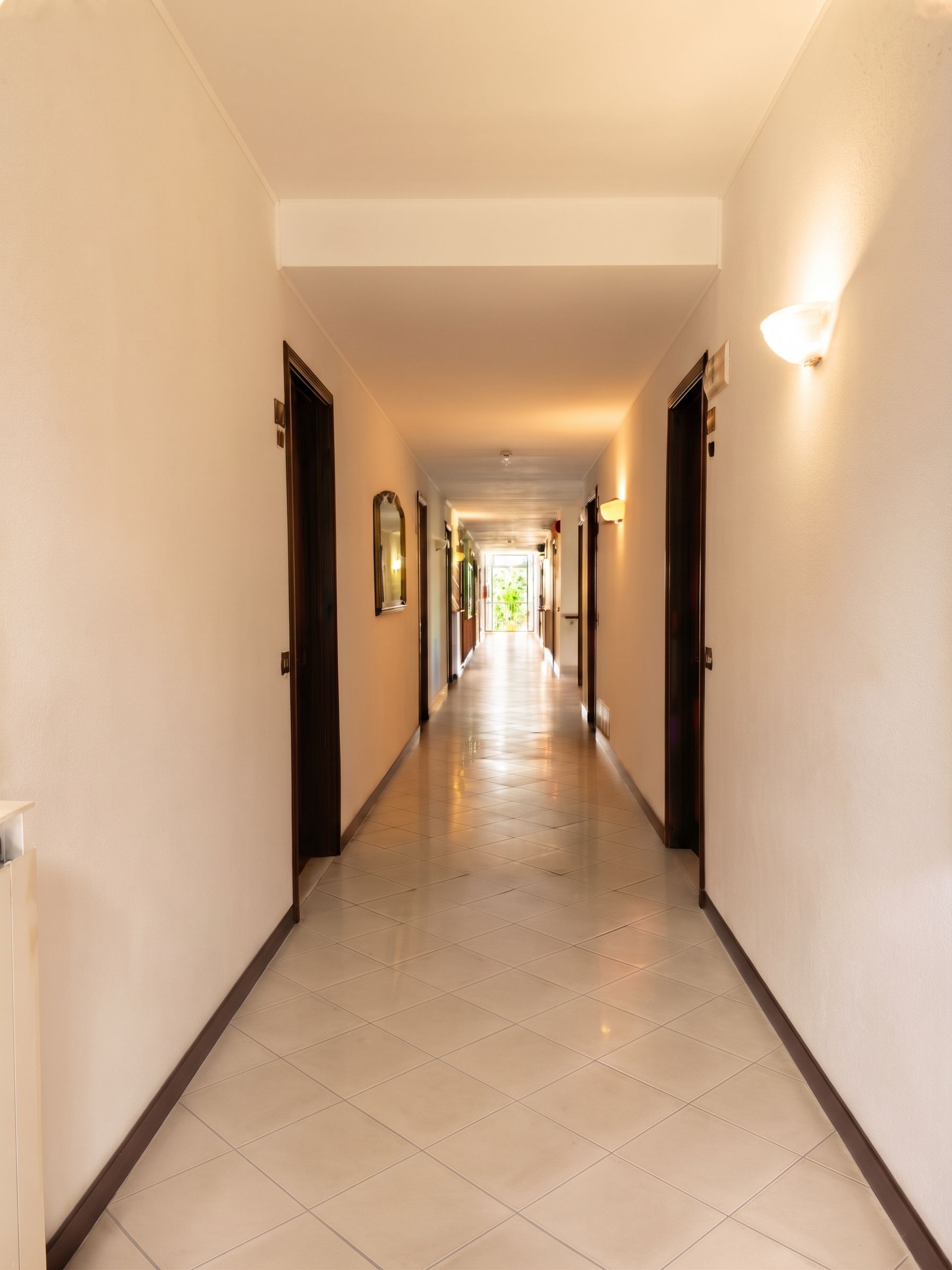 Corredor longo e estreito de hotel, com portas em ambos os lados, luminárias nas paredes e piso ladrilhado.