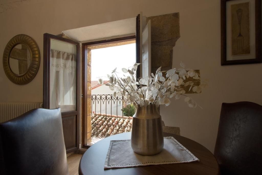 Quarto com uma mesa redonda, um vaso com flores, uma cadeira, um espelho e uma porta que dá para uma área externa.