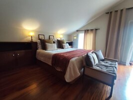 Quarto com cama grande, mesas de cabeceira, um banquinho e janela com cortinas.