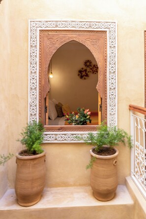 Janela com moldura branca detalhada e um espelho refletindo um quarto com uma cama e um vaso de flores.