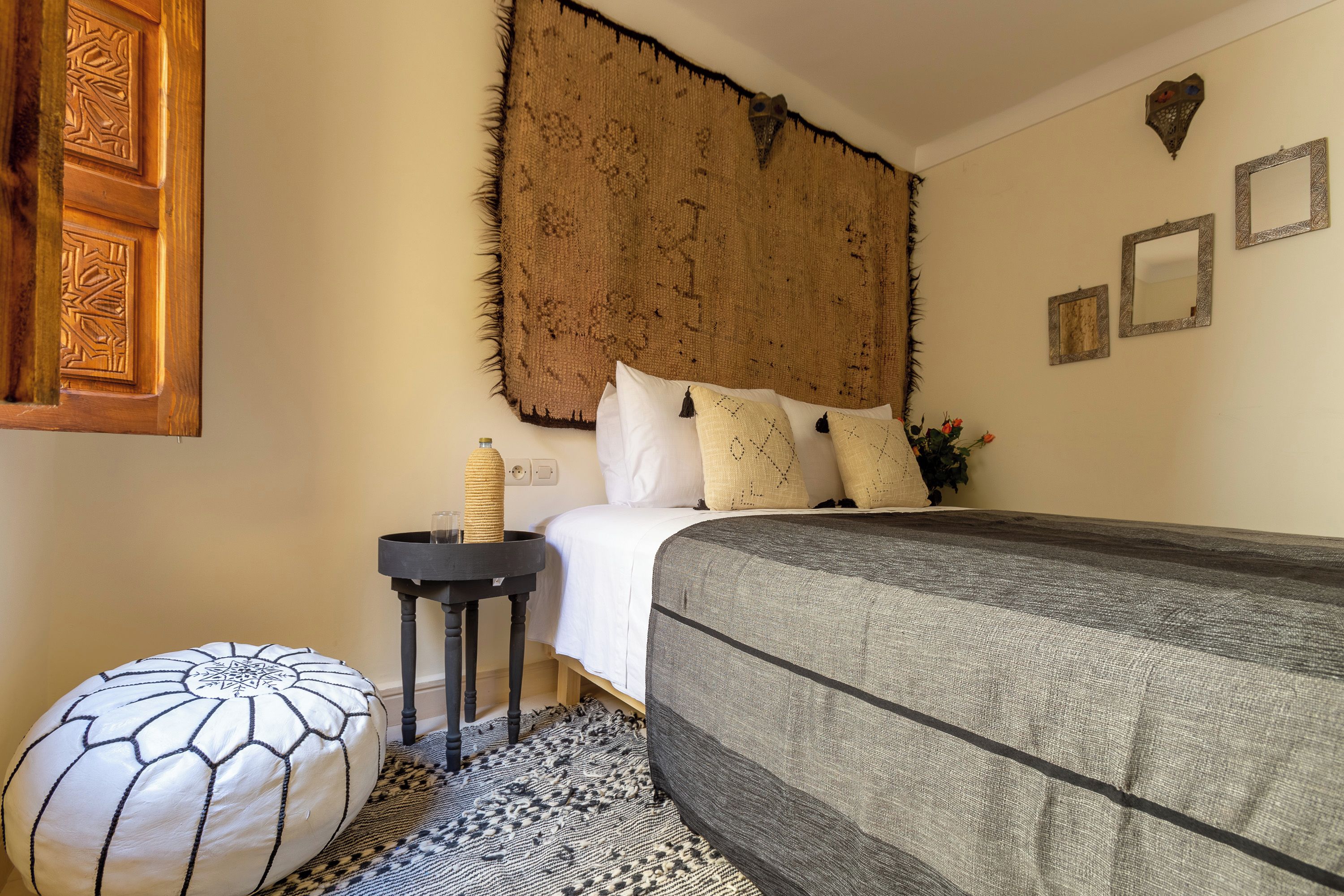 Quarto com cama, um banquinho redondo, uma mesinha com um copo e uma luminária na parede.