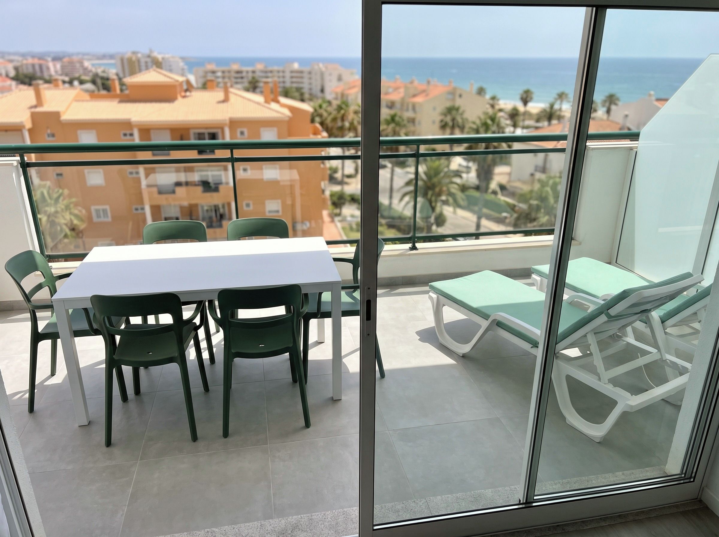 Uma varanda com mesa e cadeiras, uma espreguiçadeira e vista para o mar e edifícios.