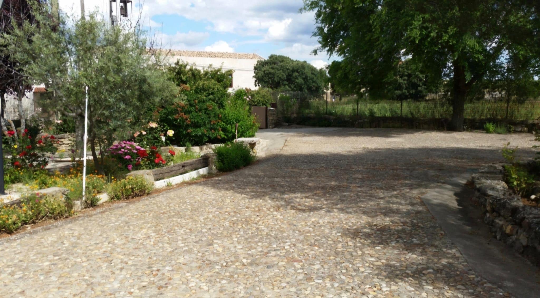 Uma entrada de cascalho leva a um edifício com fachada branca, cercado por um jardim com diversas plantas e flores.