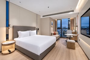 Quarto de hotel moderno com uma cama grande, escrivaninha, sofá e vista para a paisagem urbana.