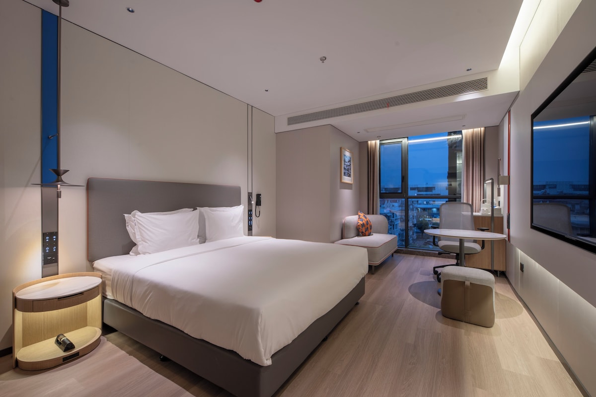 Quarto de hotel moderno com uma cama grande, uma escrivaninha e vista para a paisagem urbana.