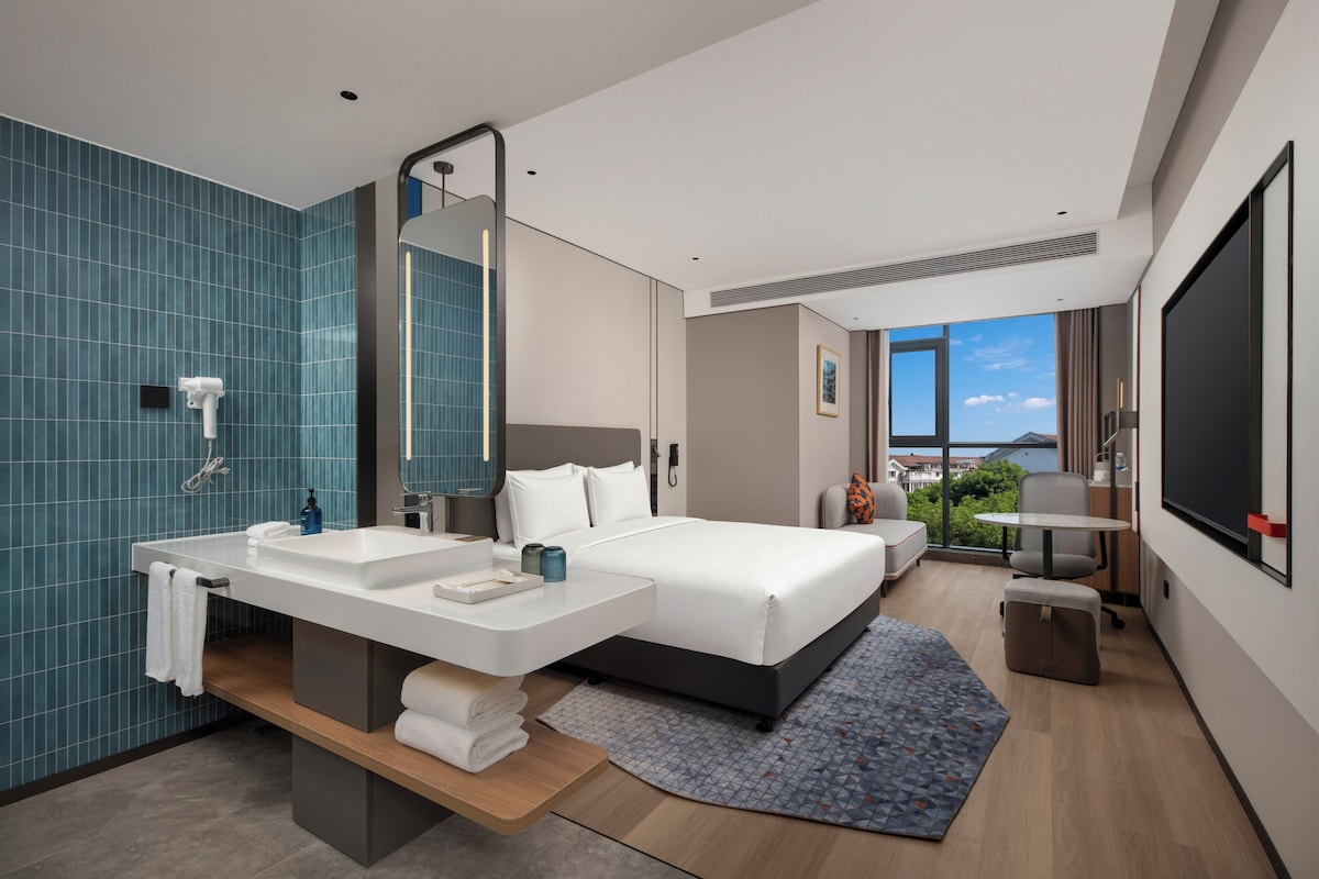 Quarto de hotel moderno com uma cama grande, área de estar separada, banheiro com um espelho amplo e vista para o exterior.