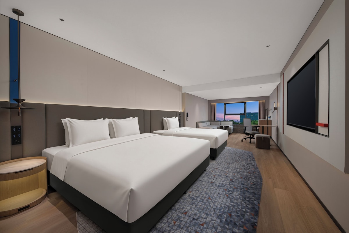 Quarto de hotel moderno com duas camas, uma escrivaninha e uma janela grande com vista para a cidade.