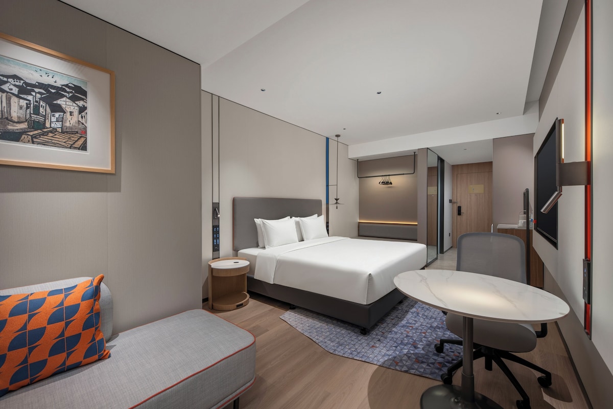 Quarto de hotel moderno com cama, escrivaninha, cadeira, mesinha de apoio e um quadro na parede.