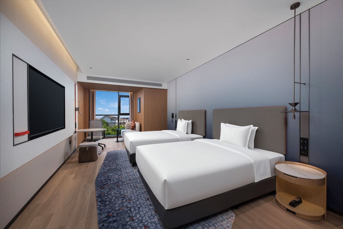 Quarto de hotel moderno com duas camas, televisão de tela plana, escrivaninha e uma grande janela com vista para a cidade.