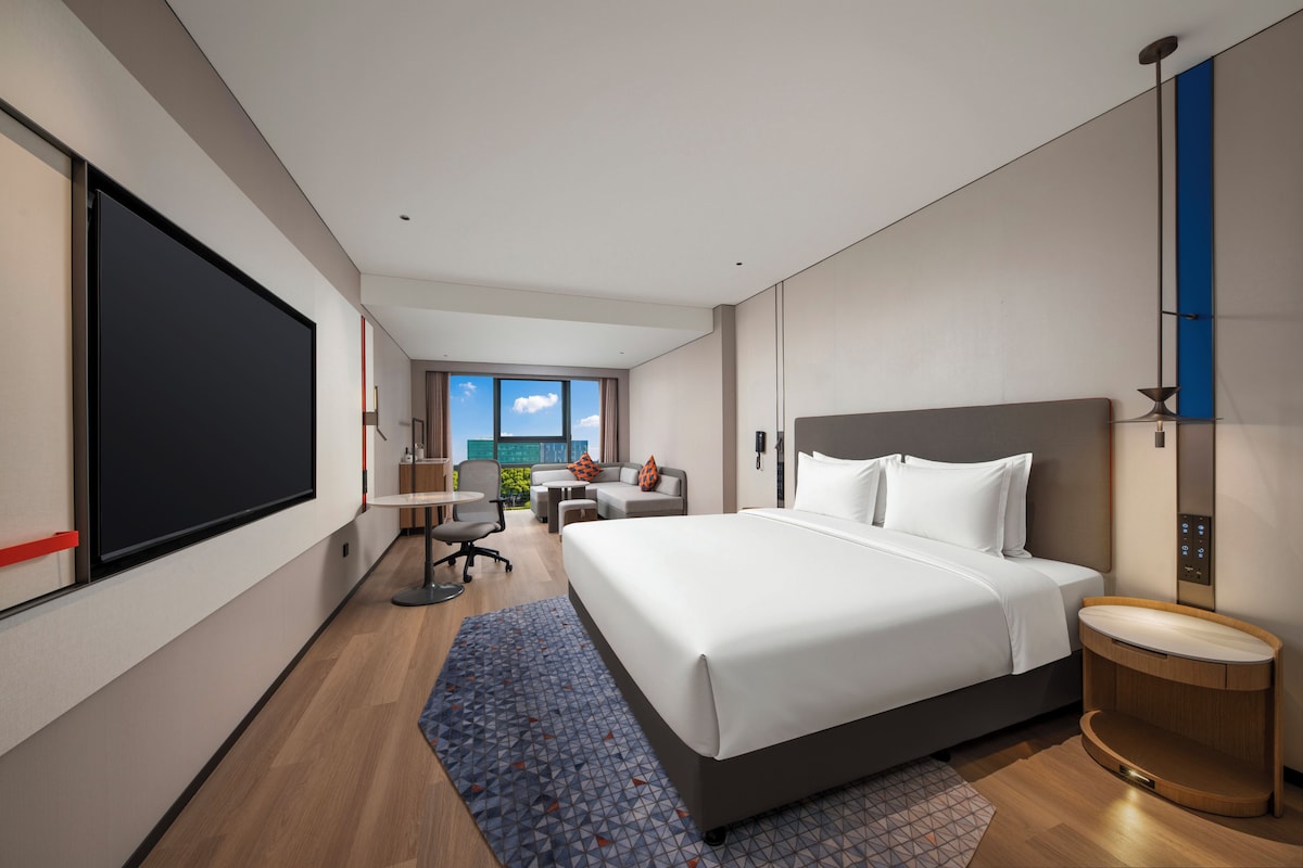 Quarto de hotel moderno com uma cama grande, uma escrivaninha, uma cadeira e vista para a cidade.