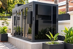Estrutura moderna e preta, retangular, com portas de vidro, possivelmente um depósito ou pequeno escritório, instalada sobre base de concreto com plantas em vasos ao redor.