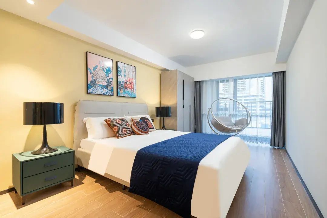 Quarto de hotel com cama, abajures, mesa de cabeceira, obras de arte na parede e vista da cidade por grandes janelas.