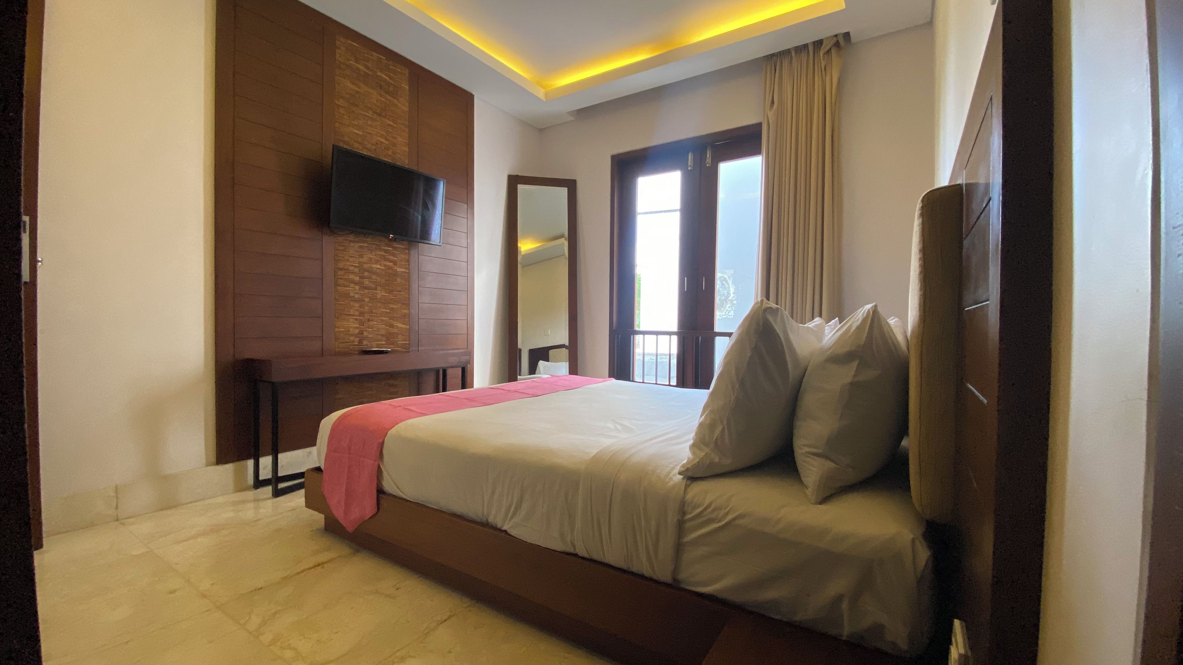 Quarto de hotel com cama, televisão de tela plana na parede, uma grande janela com cortinas e cabeceira de madeira.