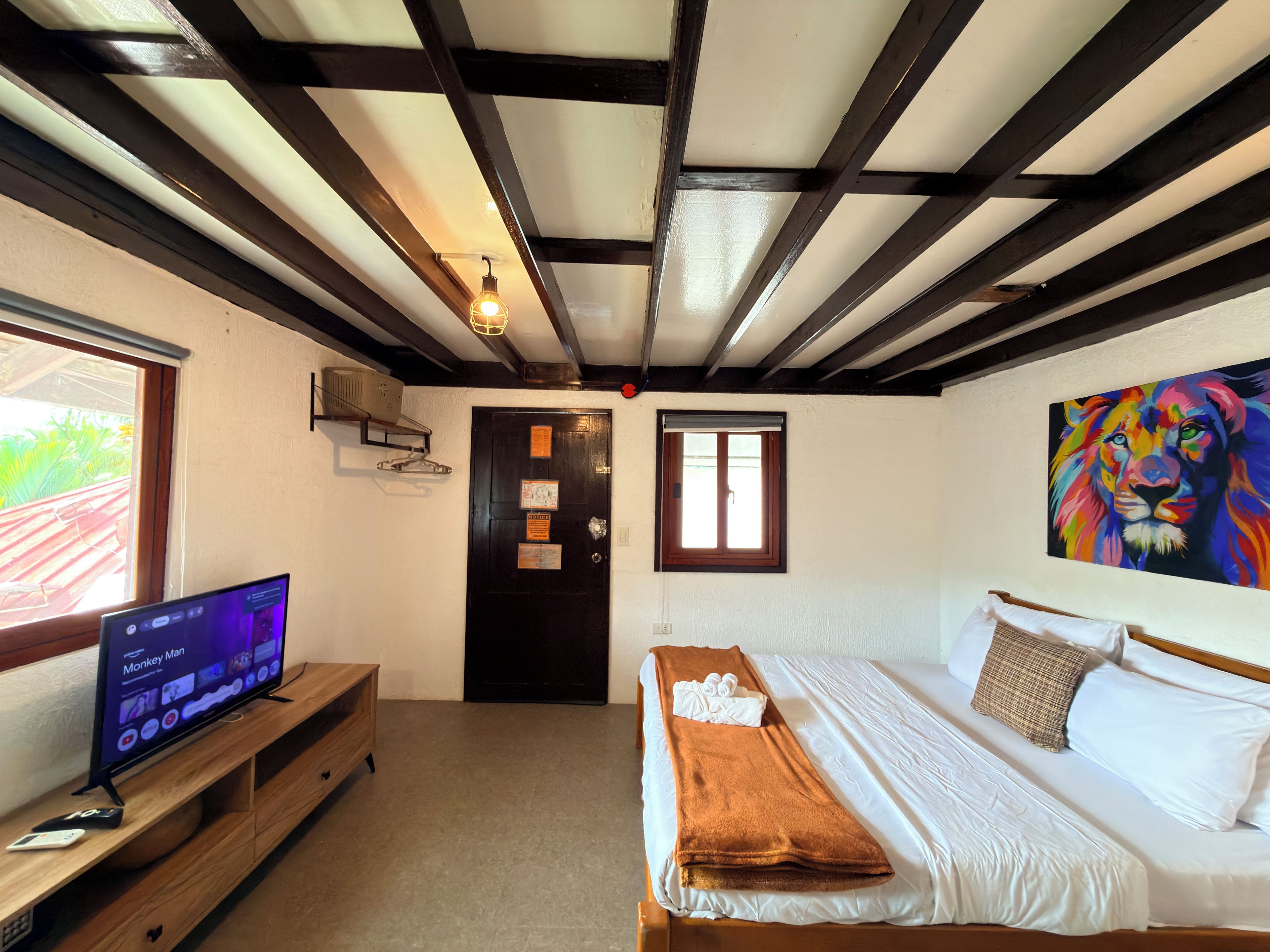 Quarto com uma cama, televisão sobre um suporte de madeira, porta com avisos, janela e um quadro na parede.