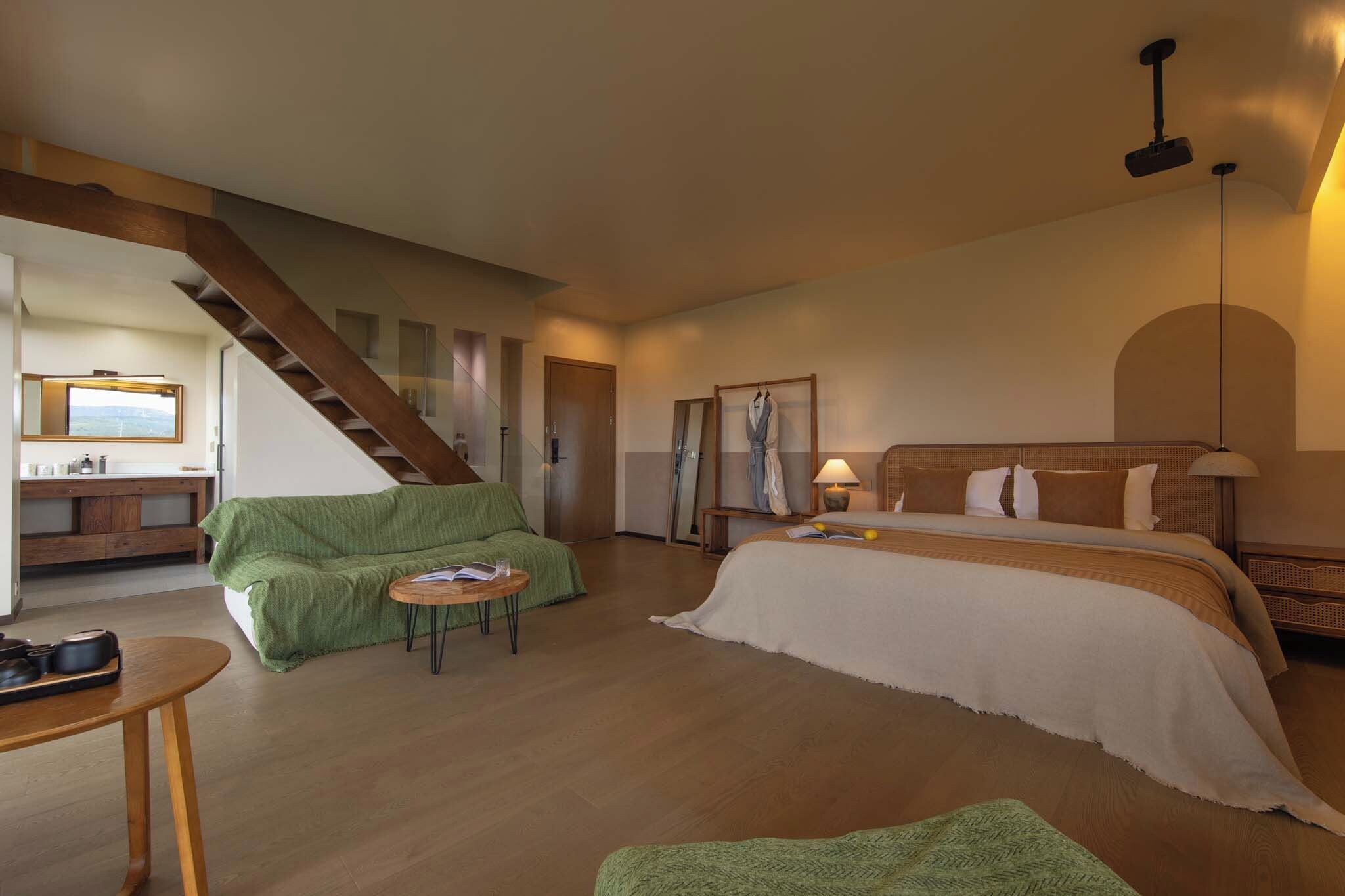 Quarto de hotel com cama, sofá verde, pequena mesa com um livro, espelho e escada.