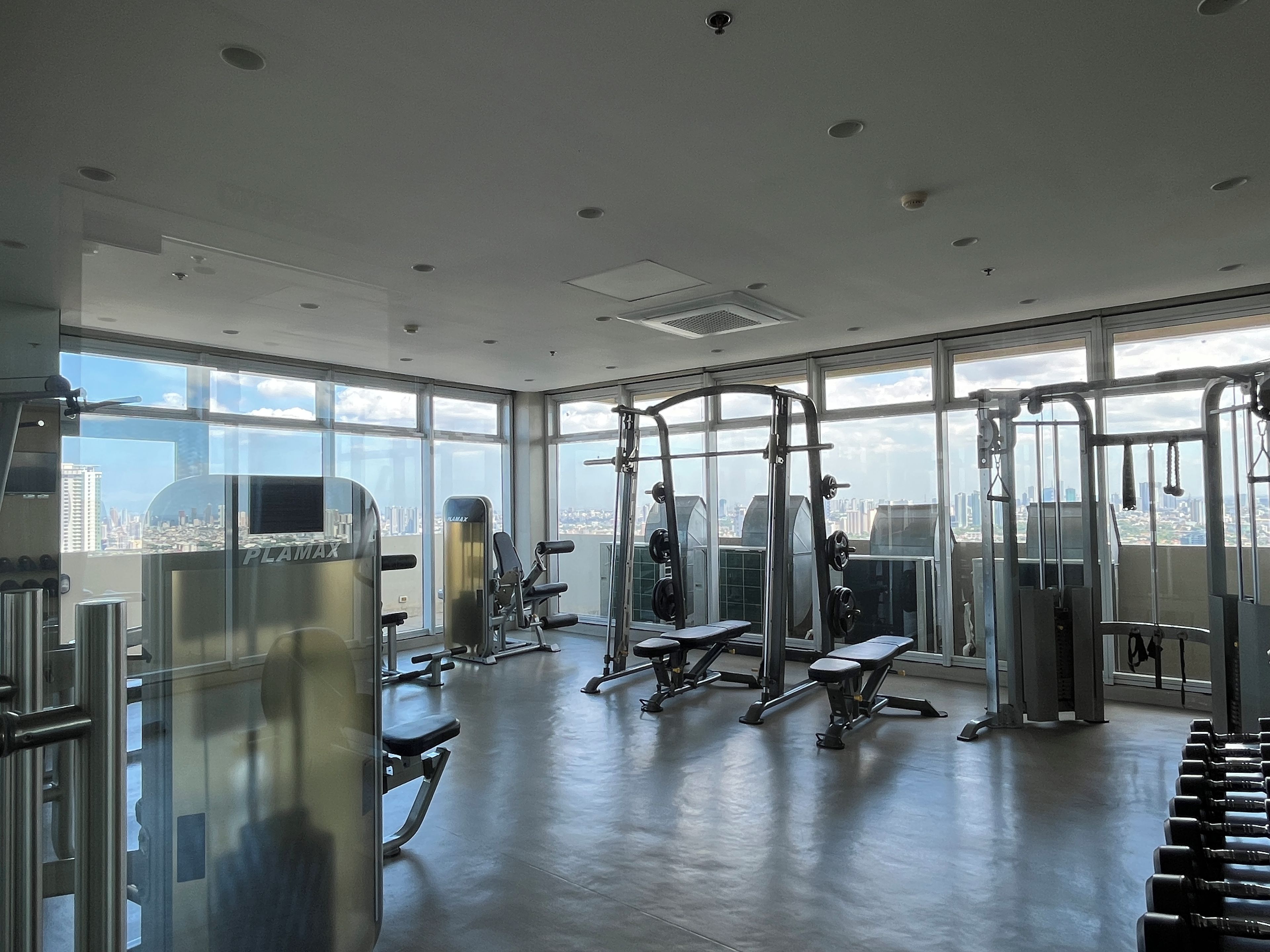 Uma academia com diversos equipamentos de ginástica, grandes janelas e vista para a cidade.