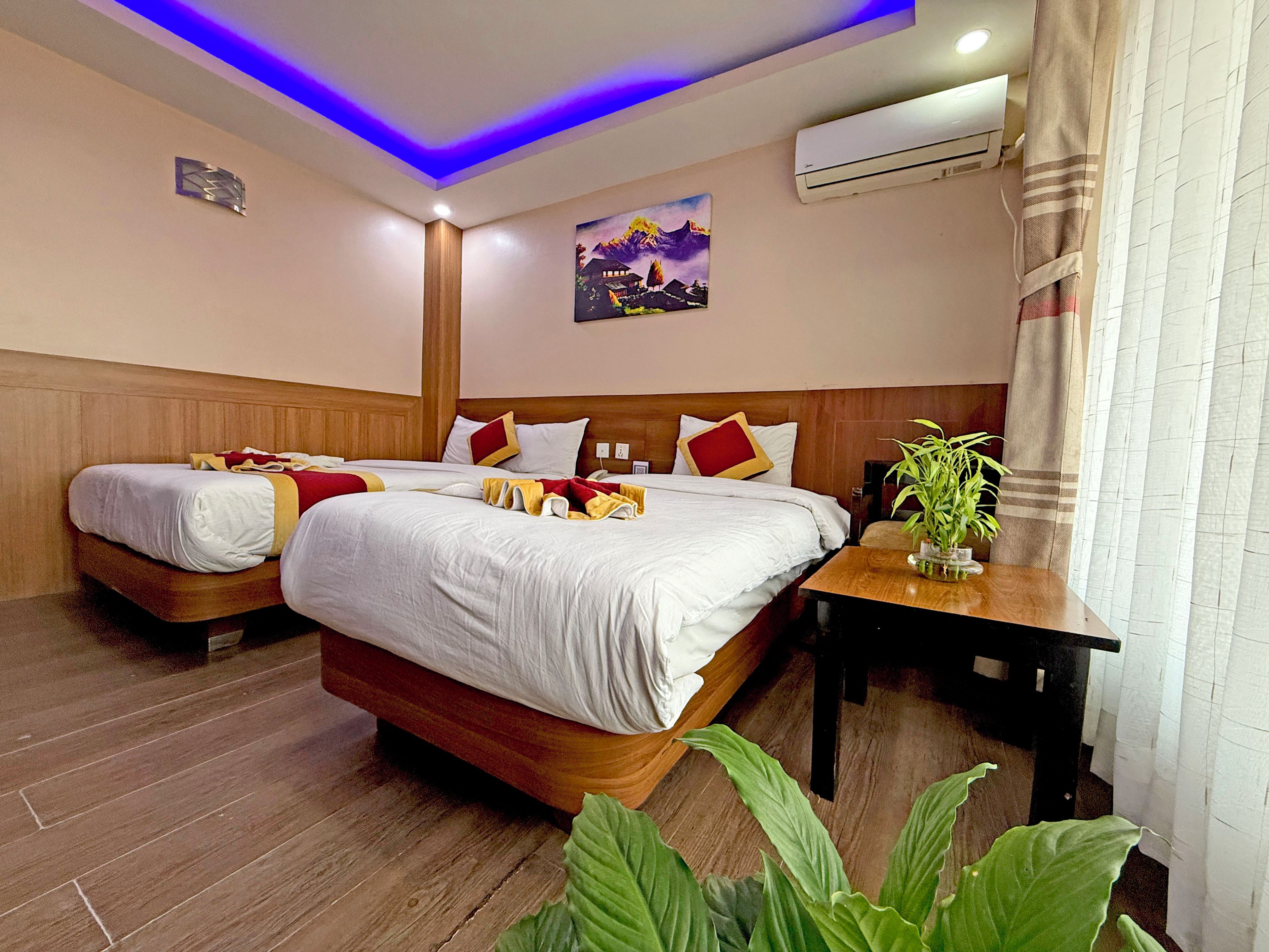 Quarto de hotel com duas camas, cabeceira de madeira, mesa de cabeceira, uma mesa e um vaso de plantas.